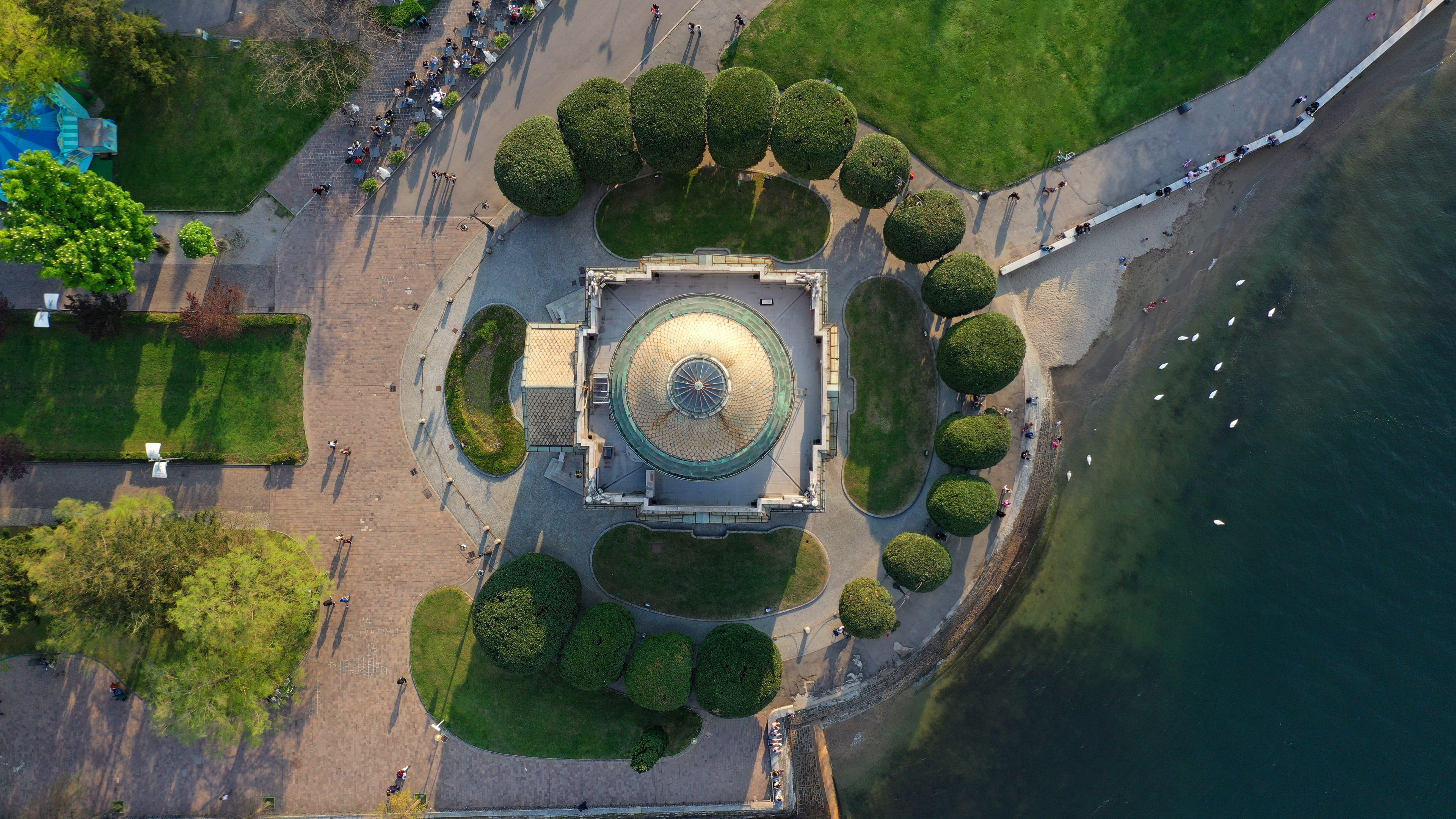 Vista aérea de um parque circular com um monumento central, ladeado por uma orla marítima e vegetação exuberante.