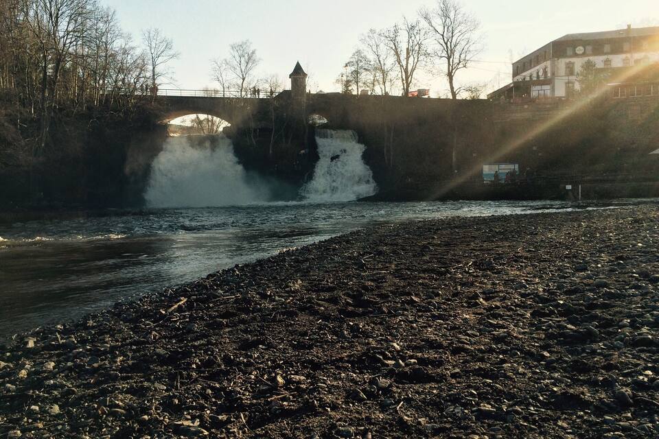 Margem rochosa de rio com uma ponte de pedra e uma cachoeira.