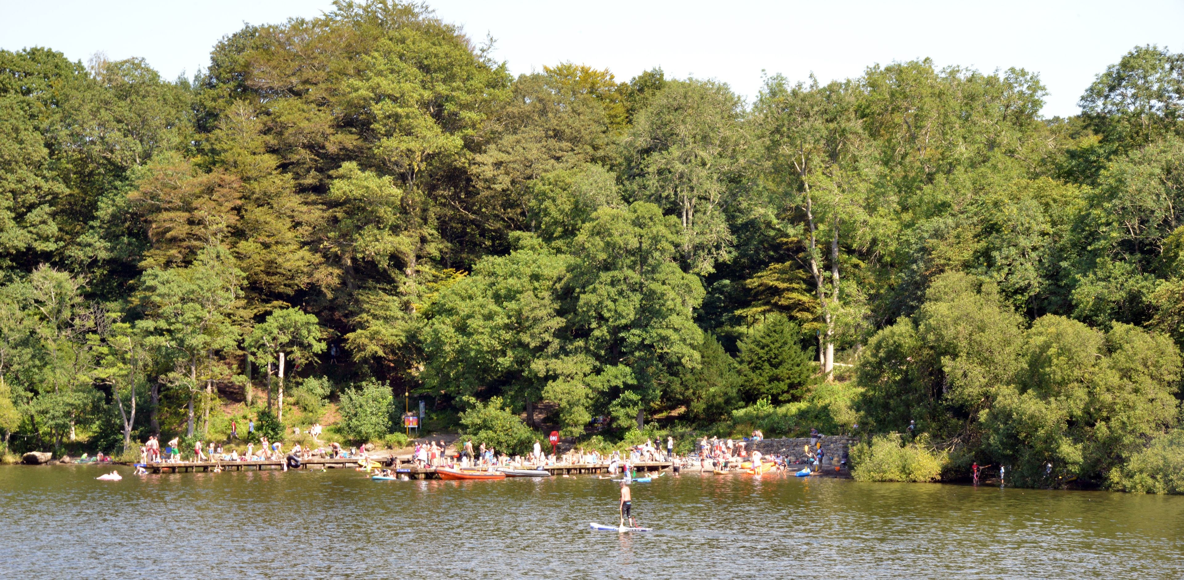 Um lago rodeado por uma floresta densa, com pessoas em um píer e uma pessoa praticando paddleboarding.
