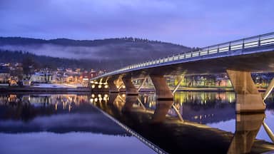 Uma ponte sobre um rio calmo que reflete as luzes de uma cidade e uma colina envolta em névoa ao fundo.