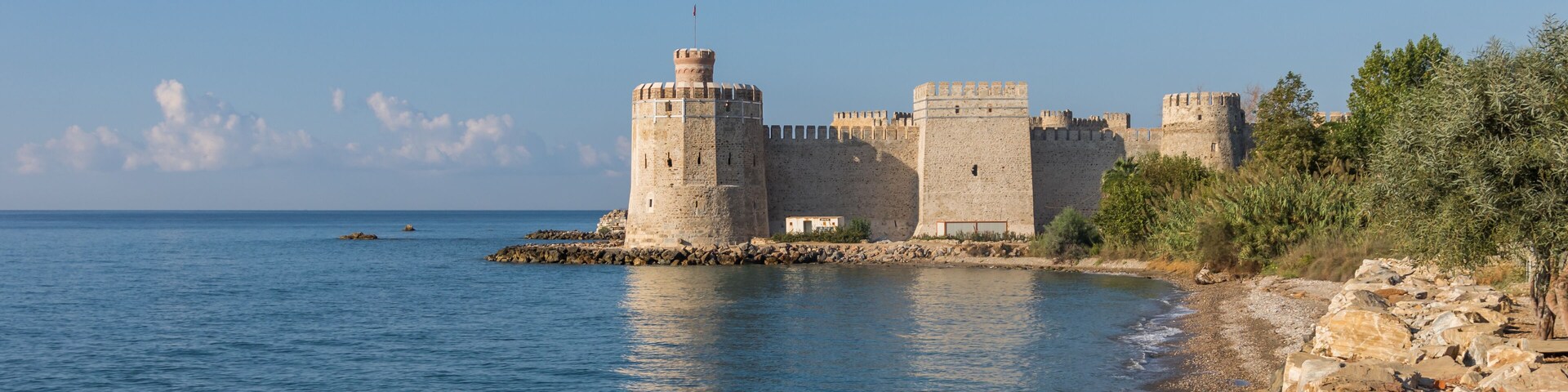 Um castelo costeiro com várias torres, rodeado por uma linha costeira rochosa e mar calmo.