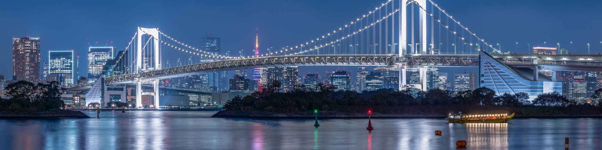 Horizonte urbano com uma ponte destacada sobre a água, edifícios iluminados e um barco a vapor.