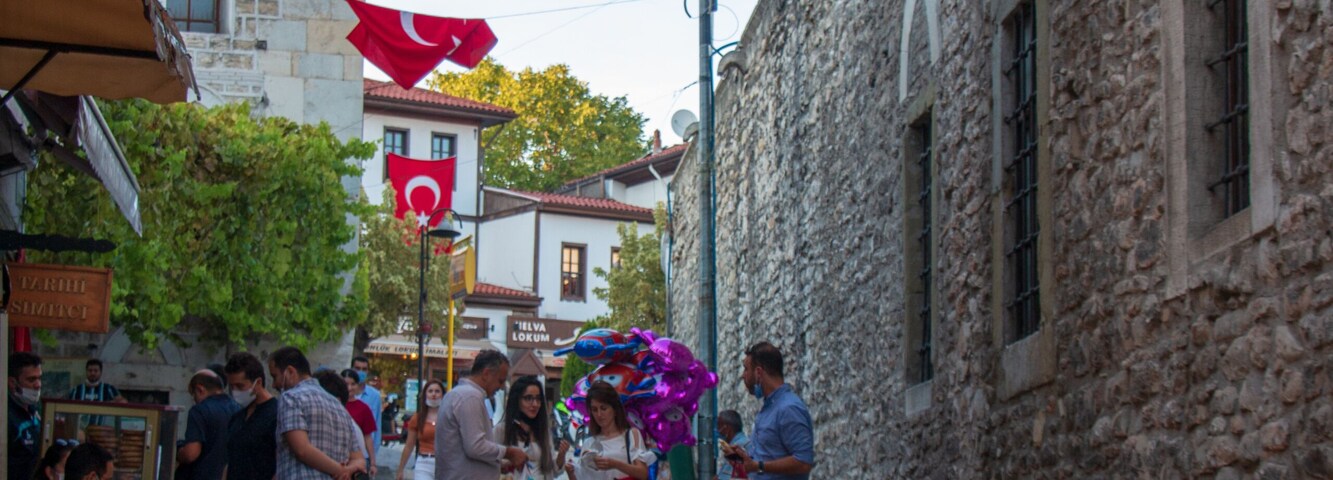 Uma rua de paralelepípedos flanqueada por edifícios de pedra, uma bandeira turca e pessoas caminhando.