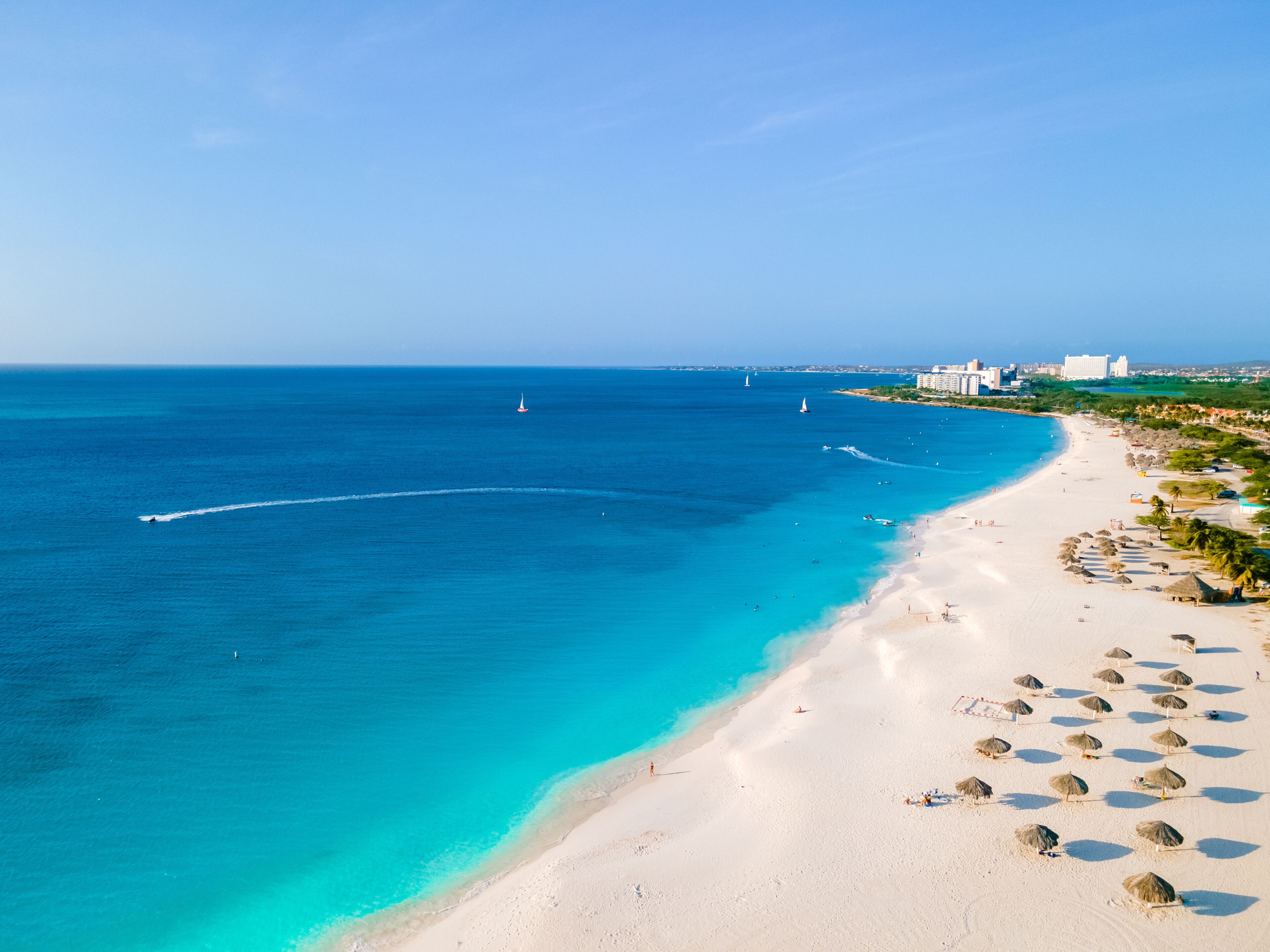 Uma praia costeira com águas turquesa cristalinas, areal arenoso e alguns veleiros ao longe.