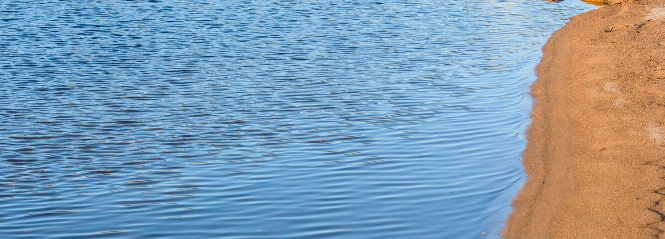 Um lago tranquilo com águas azul-claras, uma margem arenosa e uma floresta ao fundo.