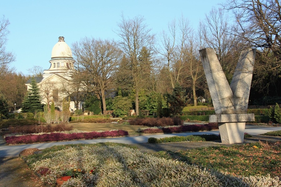 Um parque com um edifício grande e uma cúpula, uma escultura de pedra e diversos jardins ajardinados.