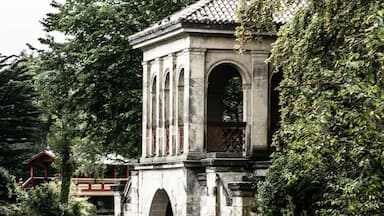 Um pavilhão de pedra com arcos e telhado cerâmico, cercado por vegetação exuberante e um corpo d'água.