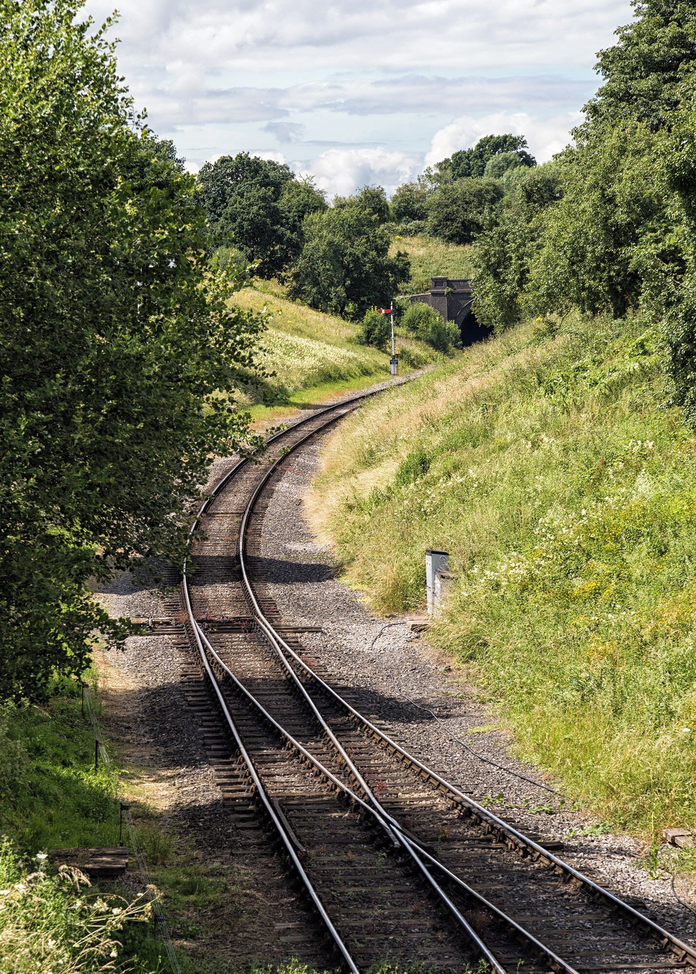 Trilhos de trem serpenteando por uma paisagem verde com árvores e uma ponte de pedra ao fundo.