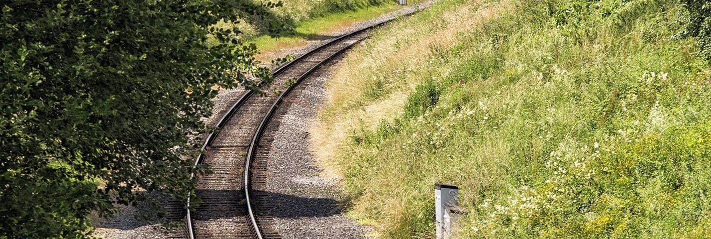 Trilhos de trem serpenteando por uma paisagem verde com árvores e uma ponte de pedra ao fundo.