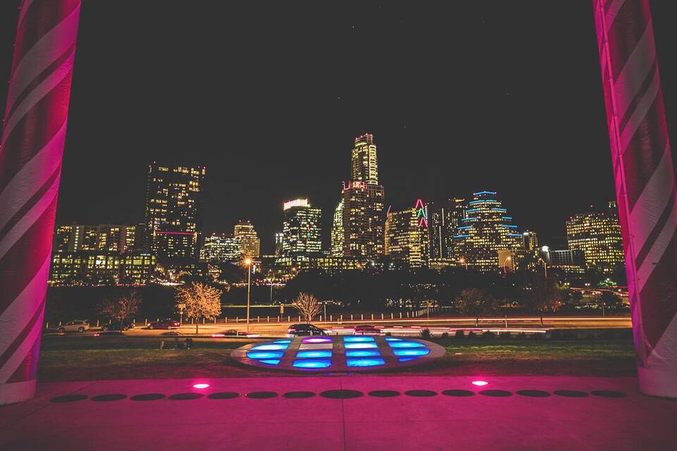 Paisagem urbana noturna com edifícios iluminados e um parque com jardineiras iluminadas em azul.