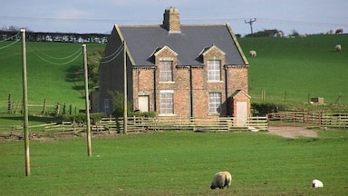Uma casa de tijolos com telhado cinzento, cercada por um campo verde e uma cerca de madeira.