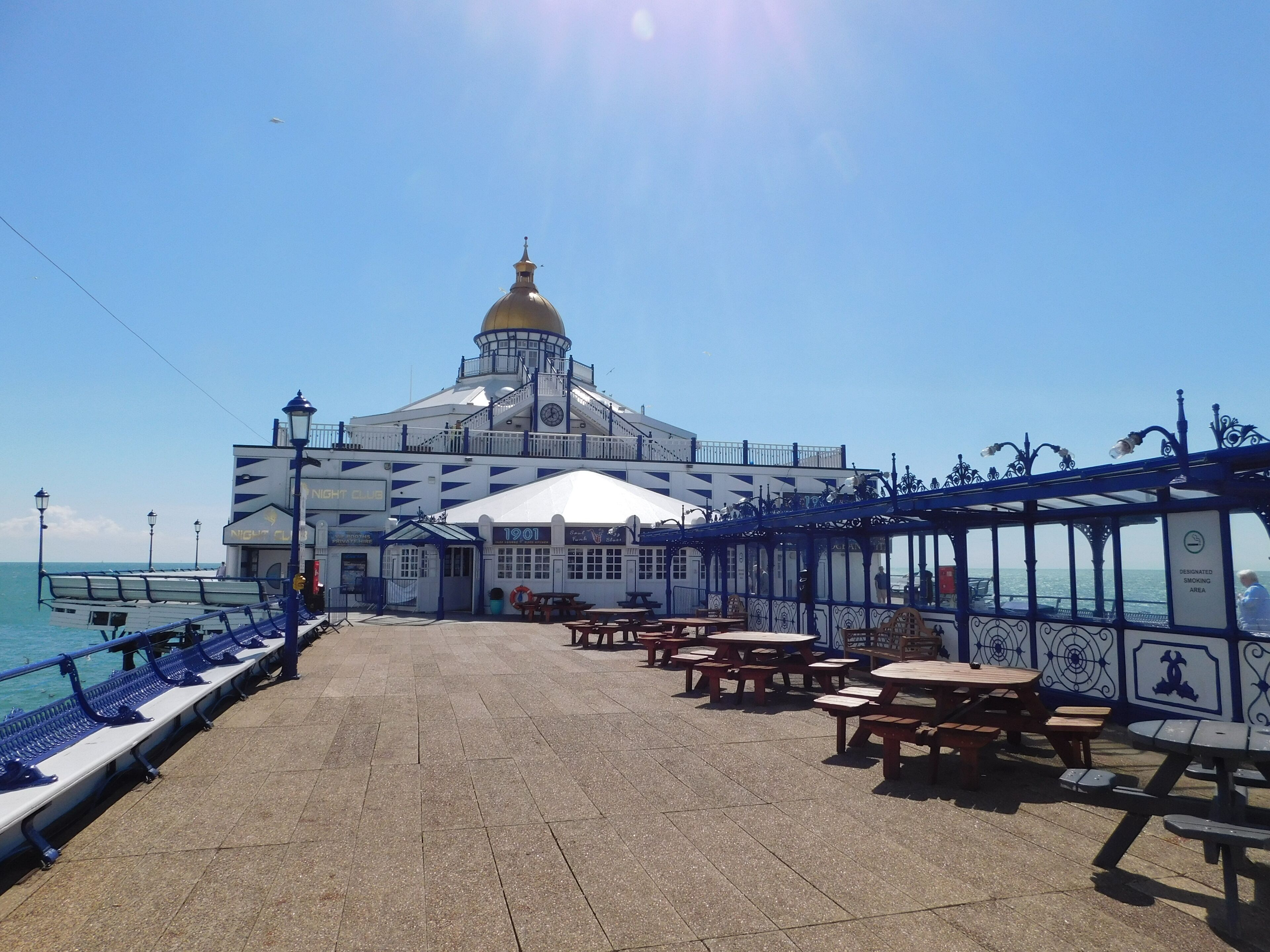 Um calçadão à beira-mar com um grande edifício branco, com cúpula dourada, bancos e guarda-corpo com vista para o oceano.