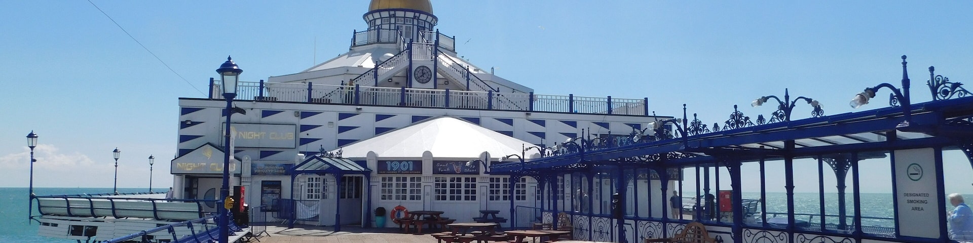 Um calçadão à beira-mar com um grande edifício branco, com cúpula dourada, bancos e guarda-corpo com vista para o oceano.