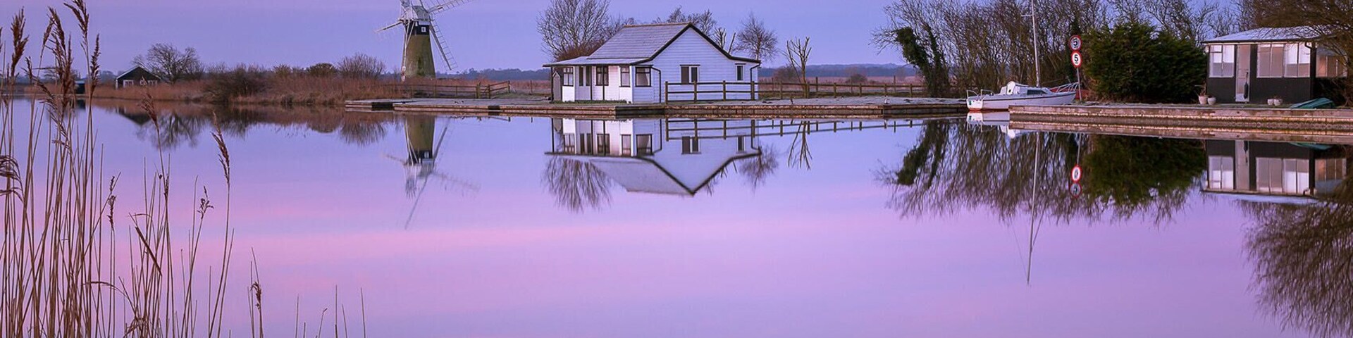 Um lago calmo com uma casa e um moinho de vento ao fundo, rodeado por juncos e árvores.