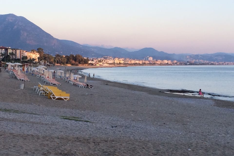 Uma praia com espreguiçadeiras e guarda-sóis, mar calmo e paisagem urbana ao fundo, com montanhas.