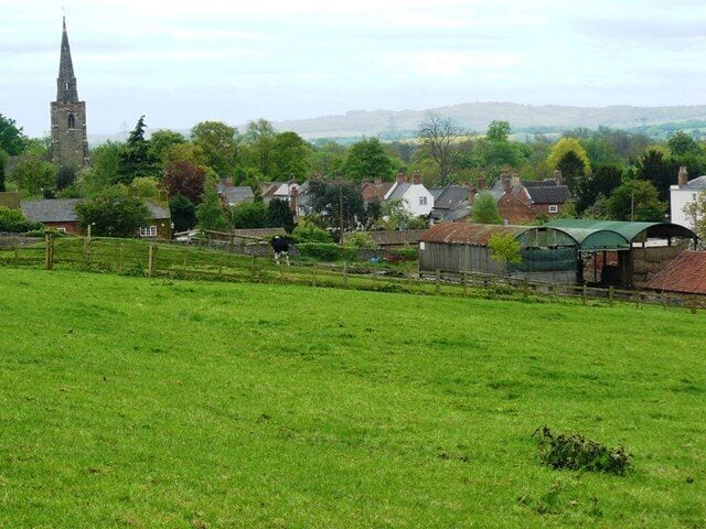 Paisagem rural com o campanário de uma igreja, campos verdes e edifícios residenciais.