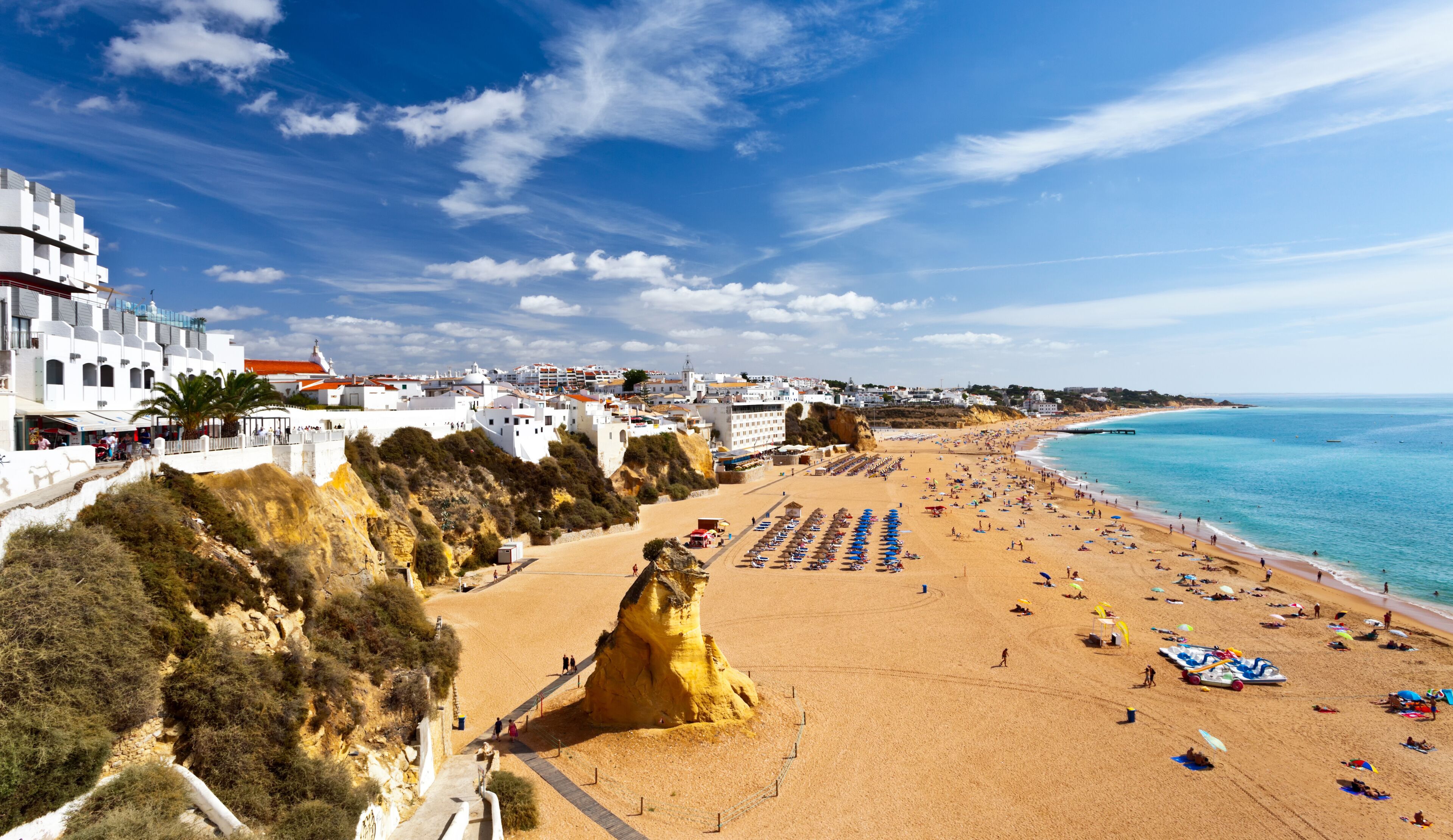 Uma vila costeira com uma praia de areia, um penhasco rochoso e edifícios brancos com telhados vermelhos.