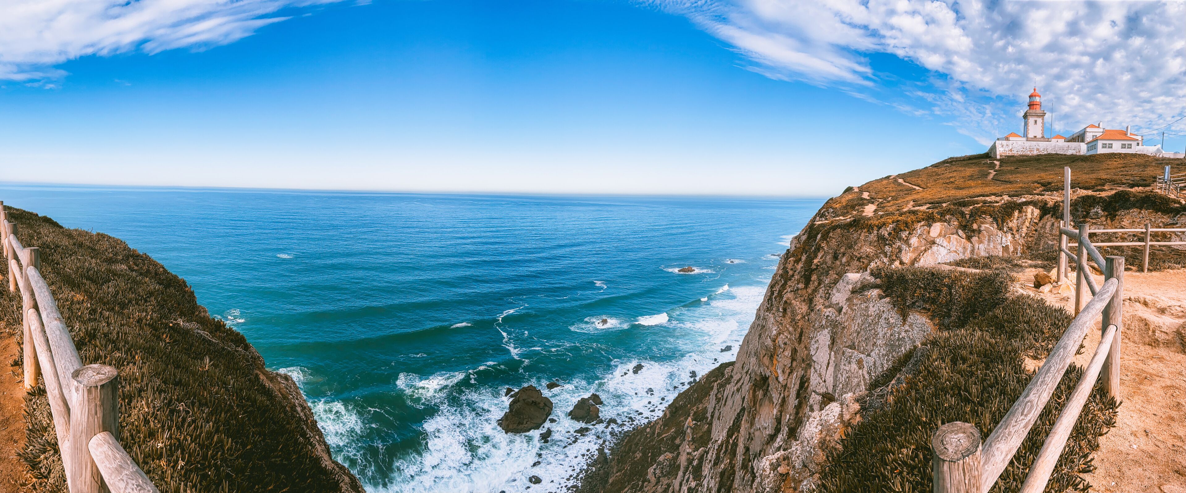 Paisagem costeira com um farol numa falésia, ondas do oceano a quebrar abaixo e um céu azul claro.