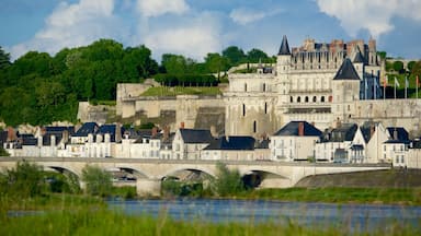 Um castelo histórico com uma ponte sobre um rio, cercado por vegetação exuberante e edifícios tradicionais.