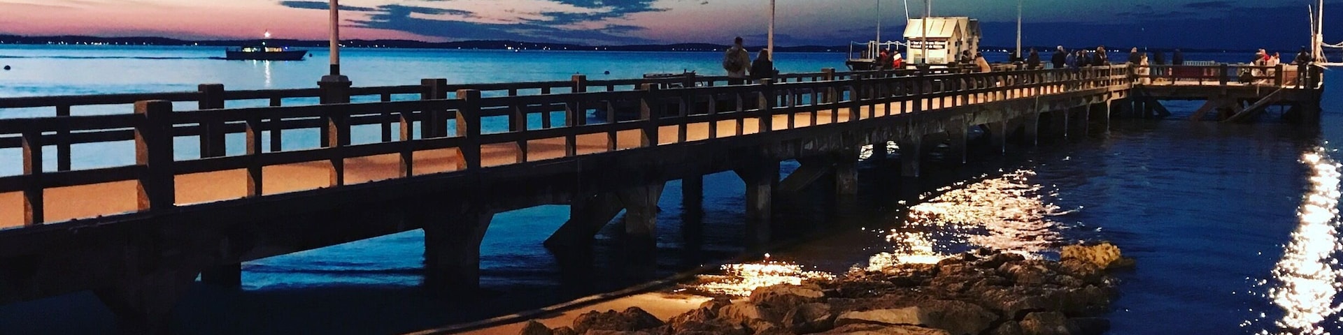 Uma praia ao entardecer, com um cais de madeira que se estende sobre a água, iluminado por postes de luz.