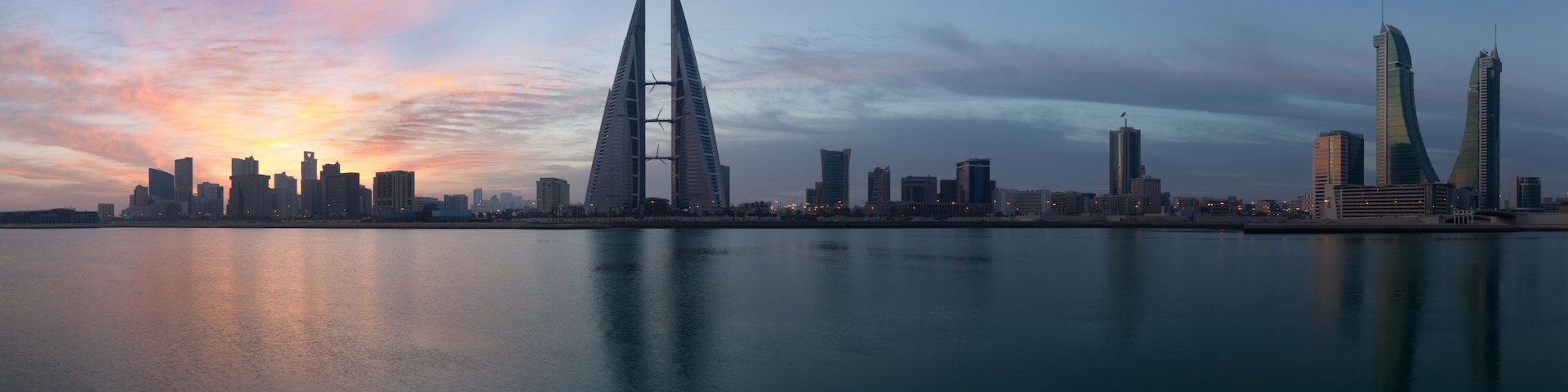 Silhueta urbana com imponentes torres gêmeas e um pôr do sol refletido em águas calmas.