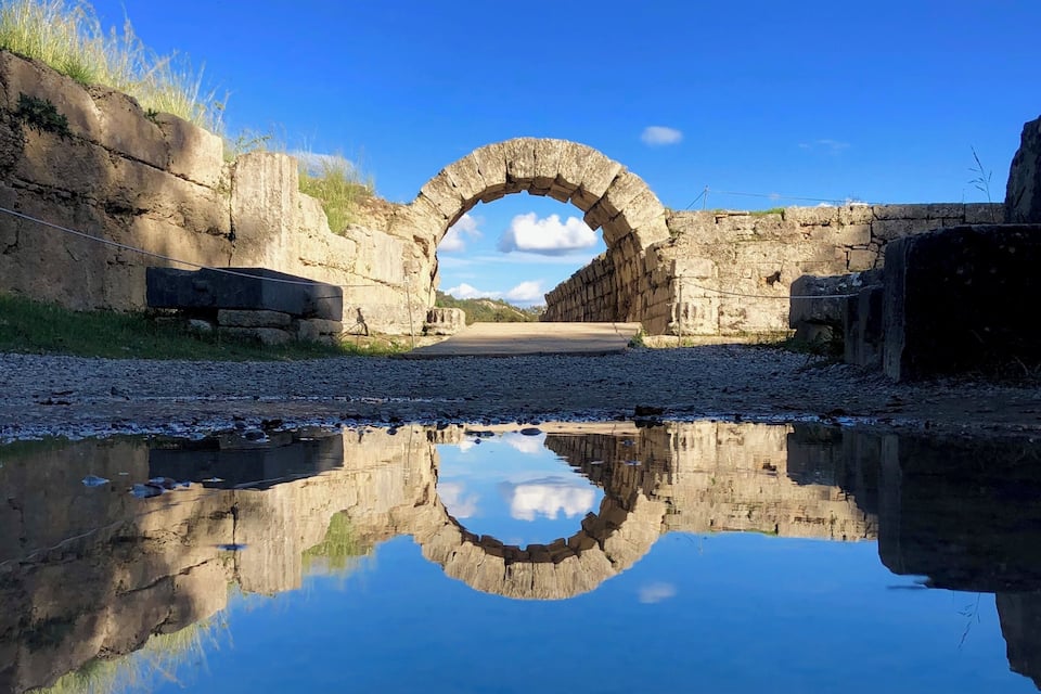 Uma ponte de pedra arqueada sobre águas calmas, refletindo o céu e as nuvens.