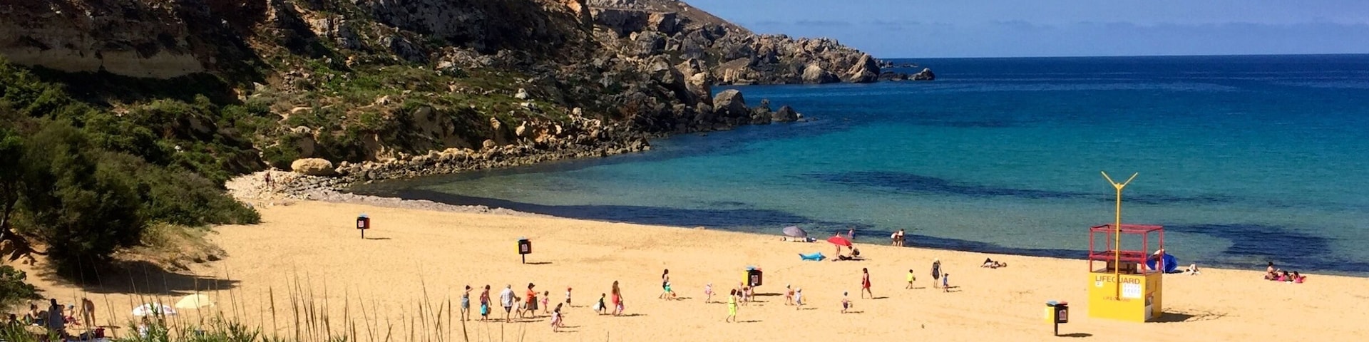 Uma praia de areia com pessoas, uma torre de salva-vidas e falésias rochosas.