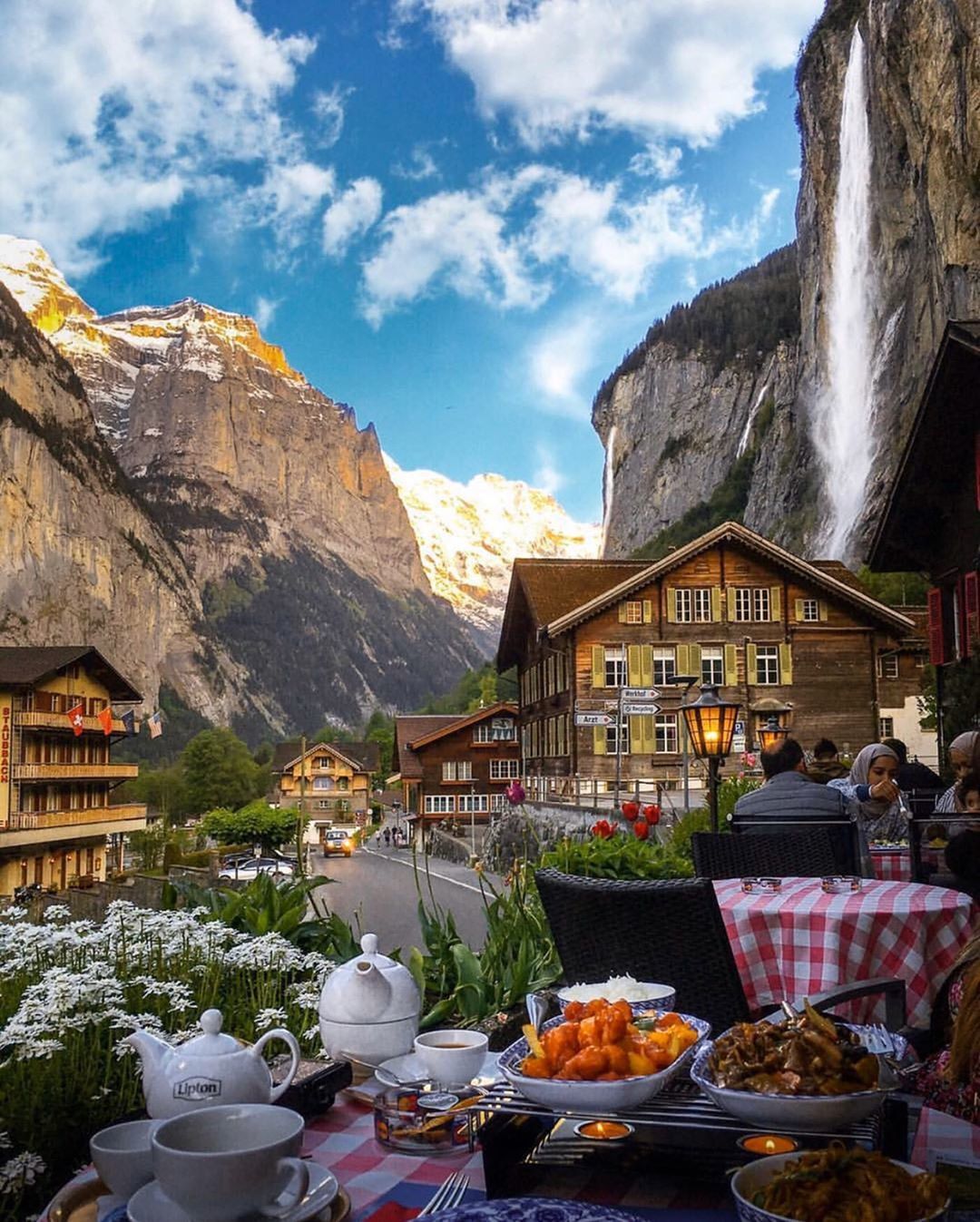 Uma aldeia de montanha com uma cascata, casas de madeira tradicionais e uma mesa de jantar posta com comida e chá.