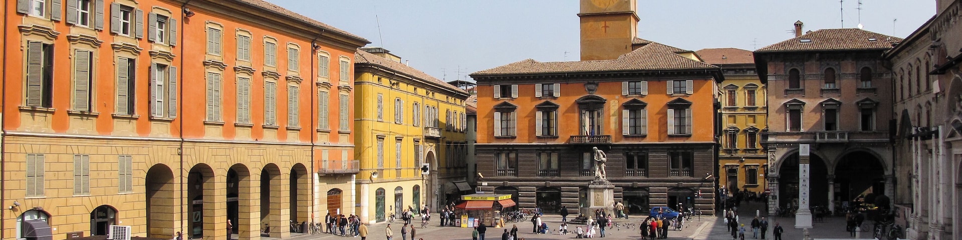 Uma praça histórica com uma torre do relógio imponente, edifícios coloridos e um espaço aberto no centro.