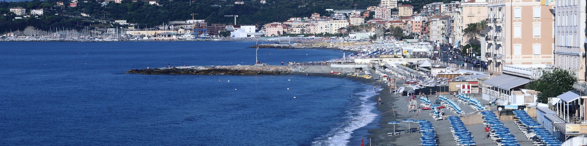 Uma cidade costeira com uma praia repleta de fileiras de guarda-sóis azuis e brancos, um píer e edifícios numa encosta.