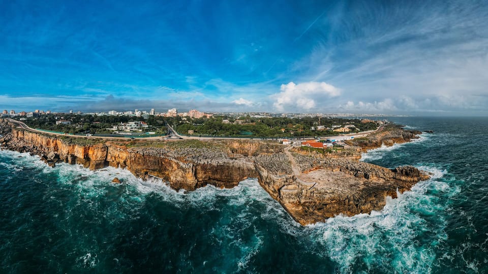 Paisagem costeira com uma península rochosa proeminente, uma ponte ligando à terra firme e um céu azul límpido.