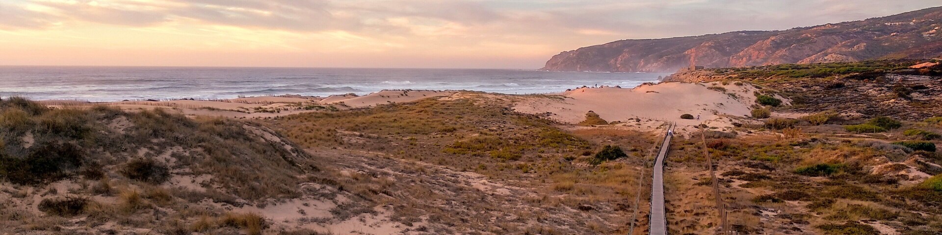 Paisagem costeira com uma passarela de madeira que leva a uma praia, cercada por dunas e falésias.