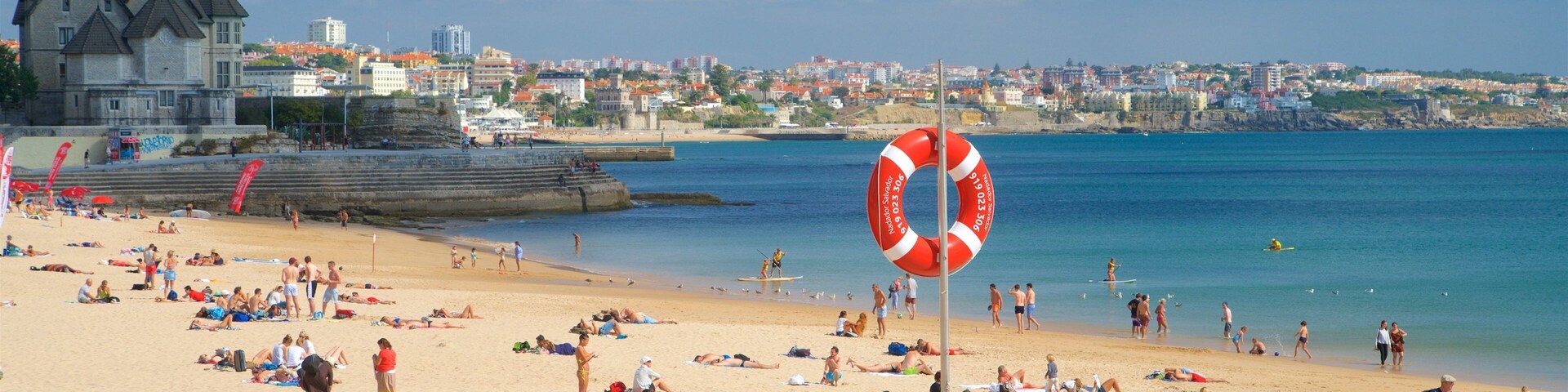 Uma praia cheia de gente, com pessoas se banhando ao sol e nadando, um flutuador salva-vidas, e um edifício histórico ao fundo.