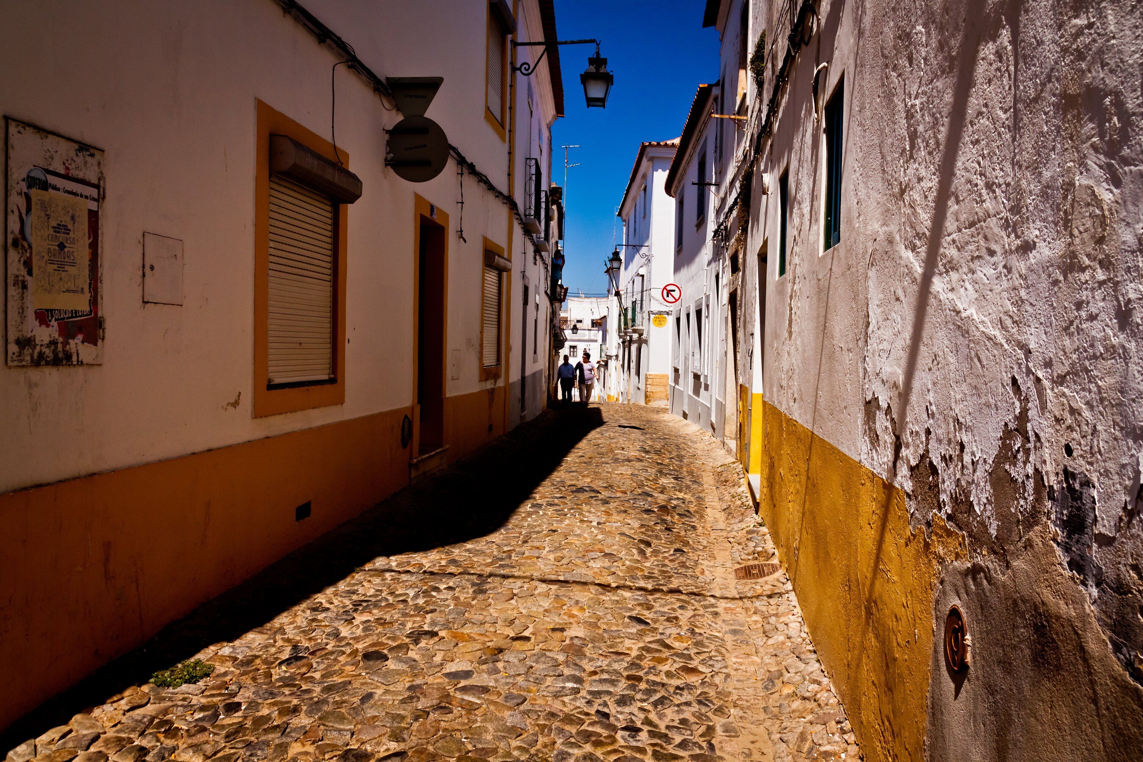 Uma rua de paralelepípedos ladeada por edifícios com fachadas amarelas e brancas, chão de pedra e céu azul claro.