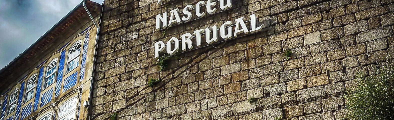 Um muro de pedra com a inscrição "AQUI NASCEU PORTUGAL" e um edifício com janelas de padrão azul e branco.