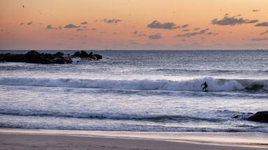 Uma praia com ondas e um surfista ao pôr do sol.