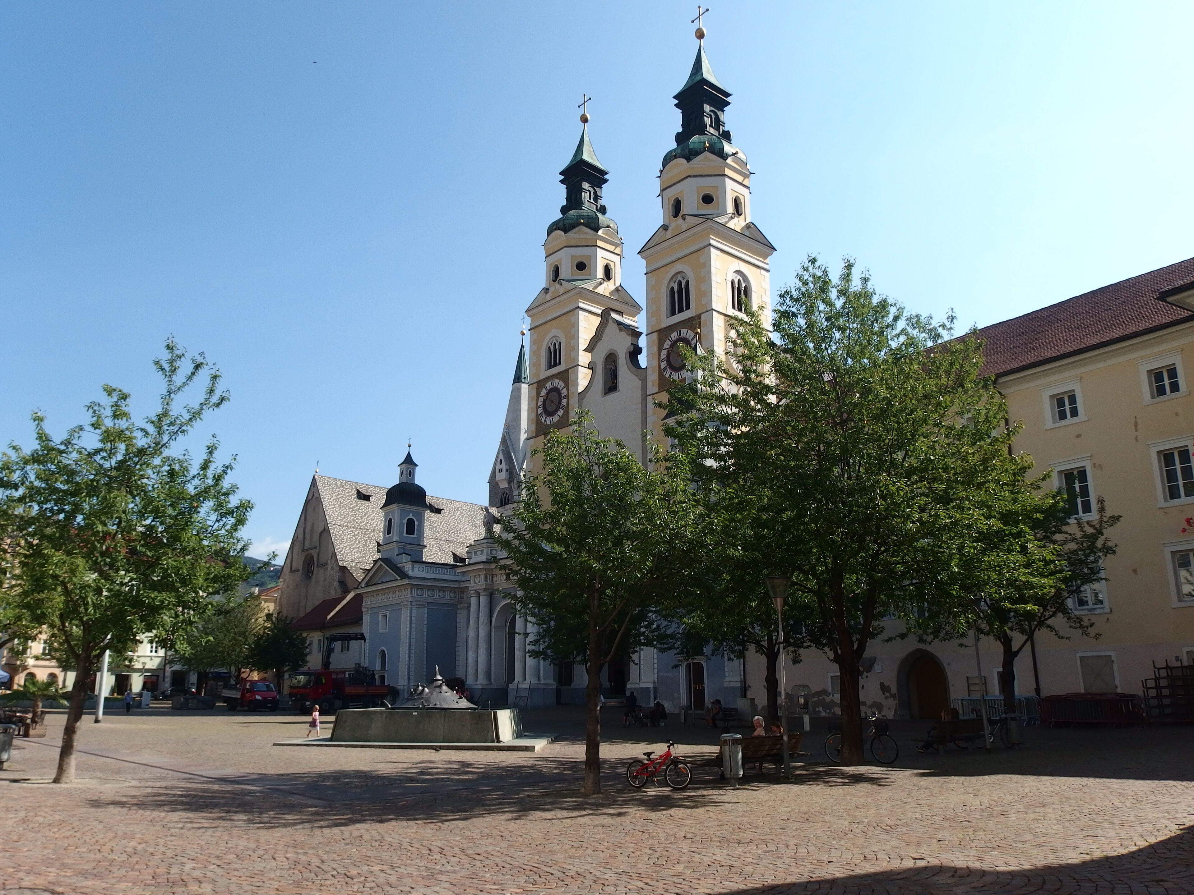 Uma igreja histórica com duas torres e uma fonte numa praça de calçada de pedra.