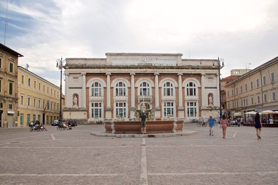Um edifício histórico com uma fonte central e estátuas, rodeado por ruas de calçada e outros edifícios.