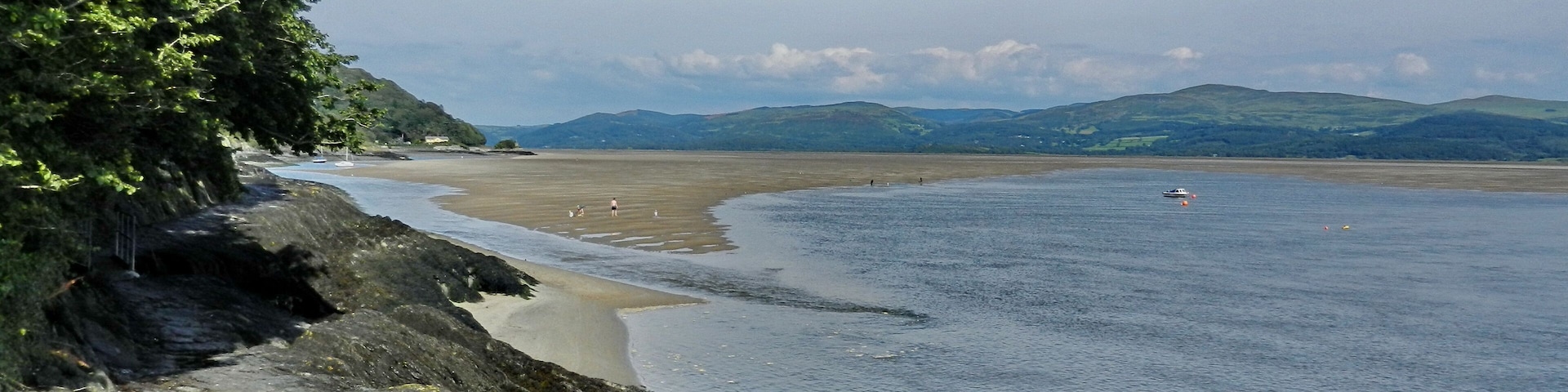 Paisagem costeira com costas rochosas, uma praia de areia e colinas ao longe, sob um céu claro.