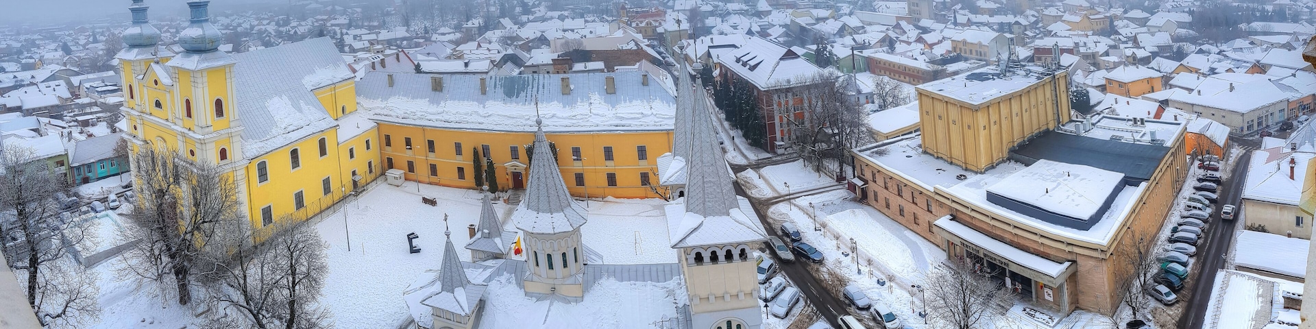 Paisagem urbana coberta de neve, com edifícios amarelos e brancos em destaque, incluindo uma igreja com campanário e um edifício amarelo grande com telhado vermelho.