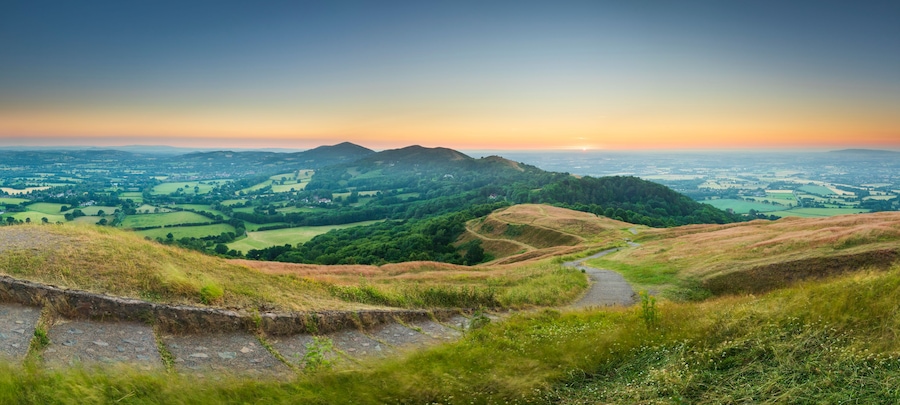 Vista panorâmica de uma paisagem montanhosa com um caminho sinuoso, campos verdes e colinas ao longe.