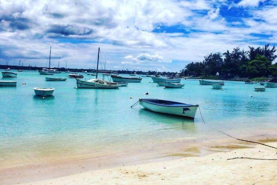 Uma praia com vários barcos pequenos ancorados em águas azul-turquesa, uma faixa de areia e uma linha de árvores ao fundo.