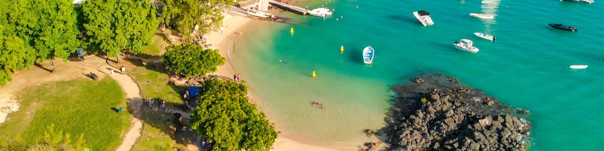 Vista aérea de uma área costeira com praia de areia, linha costeira rochosa, águas turquesa cristalinas e vários barcos.
