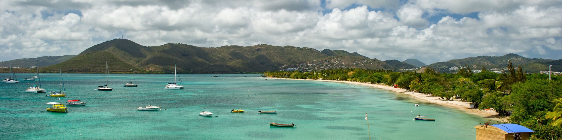 Paisagem costeira com águas azul-turquesa, barcos, uma praia arenosa e montanhas ao longe.