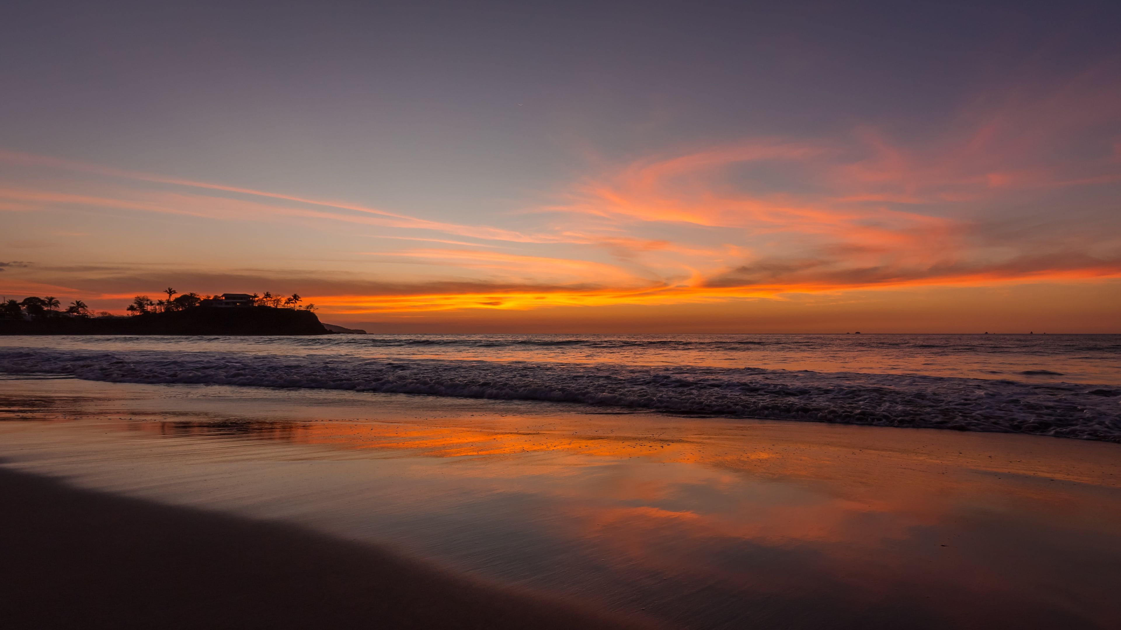 Uma praia ao pôr do sol, com ondas, uma ilha distante e céu colorido.