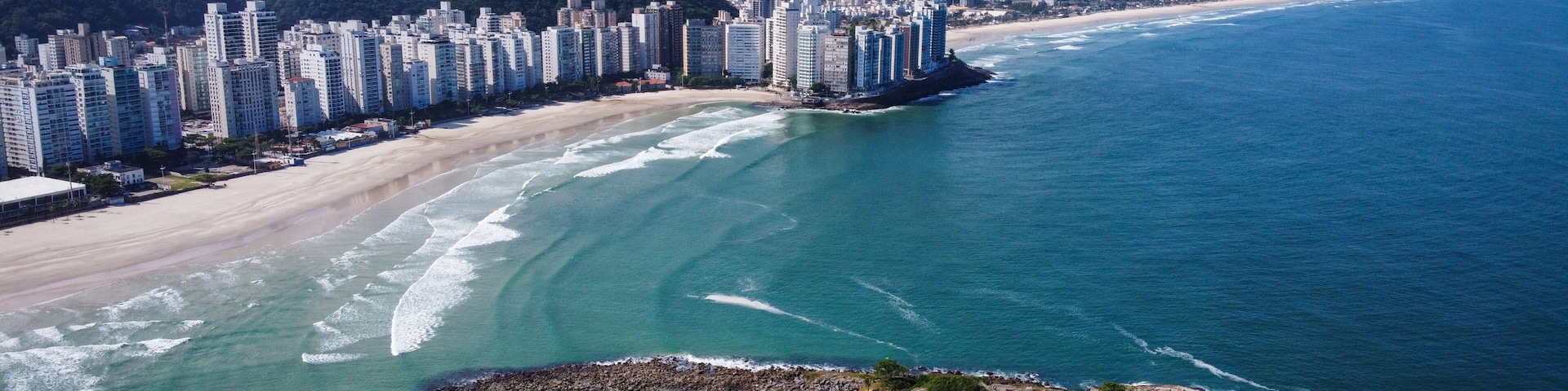 Paisagem costeira com praia, edifícios altos e um afloramento rochoso no mar.