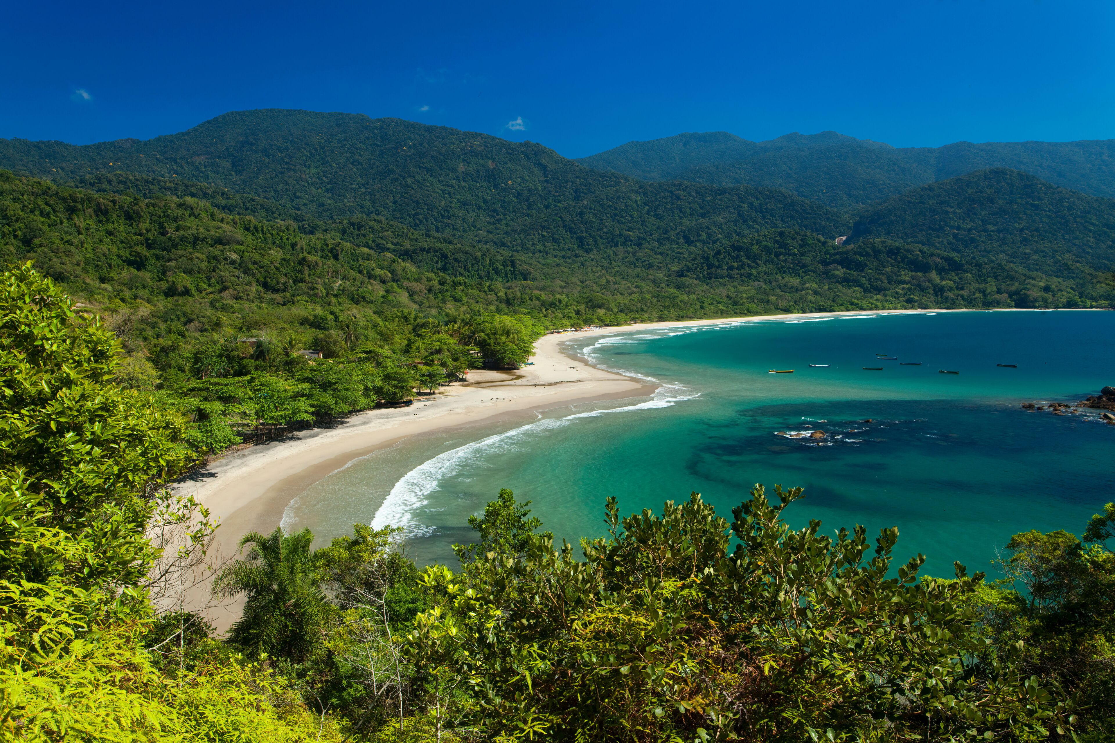 Paisagem costeira com uma praia de areia, águas azul-turquesa e montanhas verdejantes.