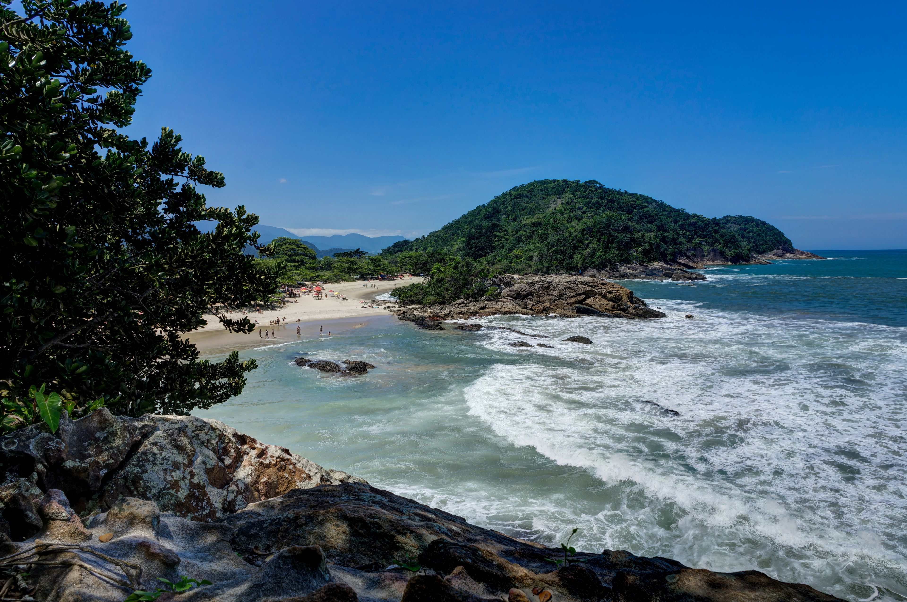 Uma praia com afloramentos rochosos, águas azul-claras e uma colina arborizada ao fundo.
