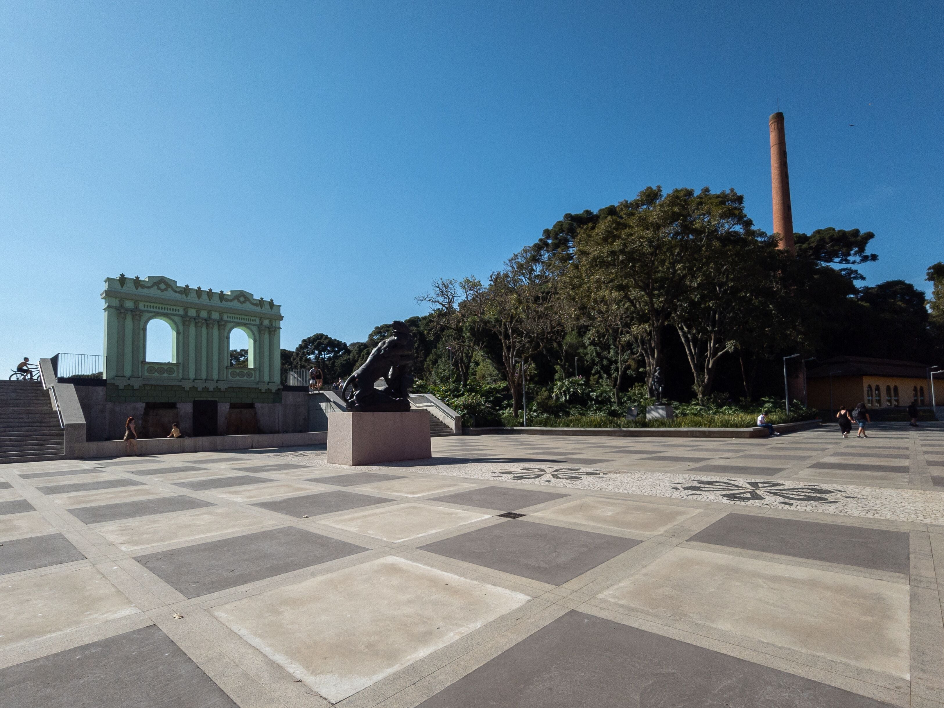 Uma praça com uma estátua, um arco verde e uma chaminé alta.