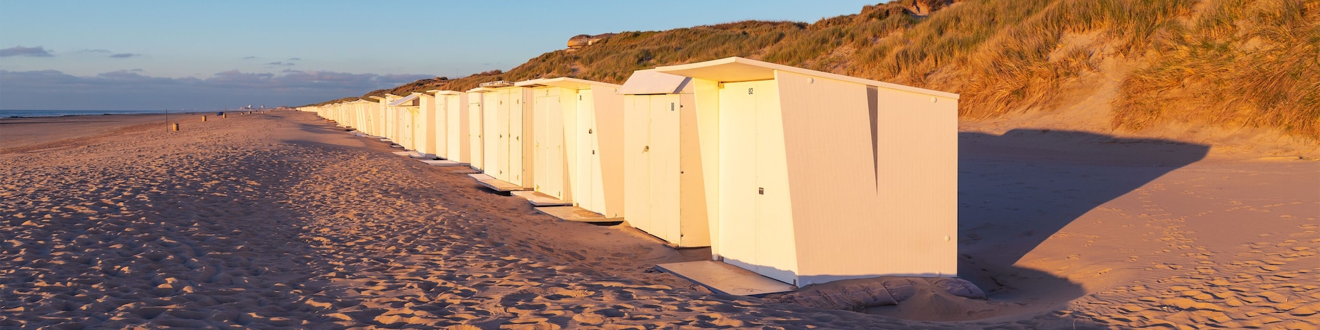 Uma praia com uma fileira de cabanas brancas à beira-mar e dunas ao fundo.